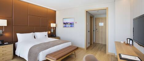 Quarto de hotel moderno com uma cama grande, mesas de cabeceira, escrivaninha com cadeira, um banco e uma televisão.