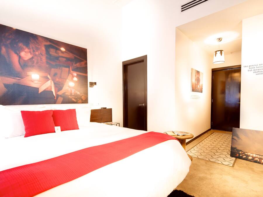 Quarto de hotel com uma cama grande, um quadro na parede, uma porta e uma mesa pequena com abajur.