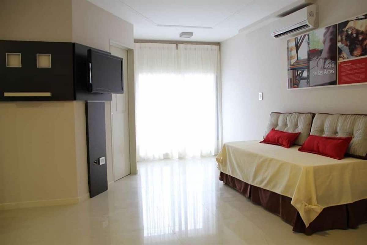 Quarto de hotel com uma cama, televisão e uma grande janela com cortinas.