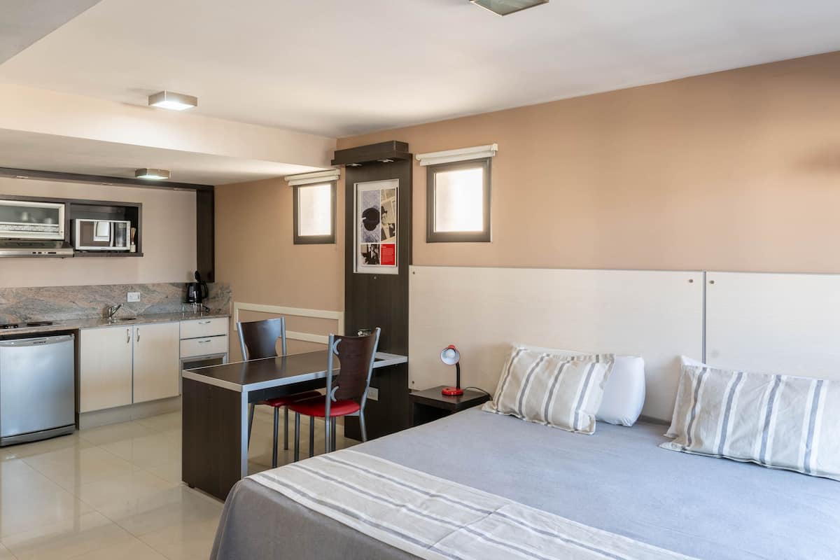 Quarto de hotel com cama, mesa de cabeceira, luminária e área de cozinha compacta, incluindo micro-ondas e cafeteira.