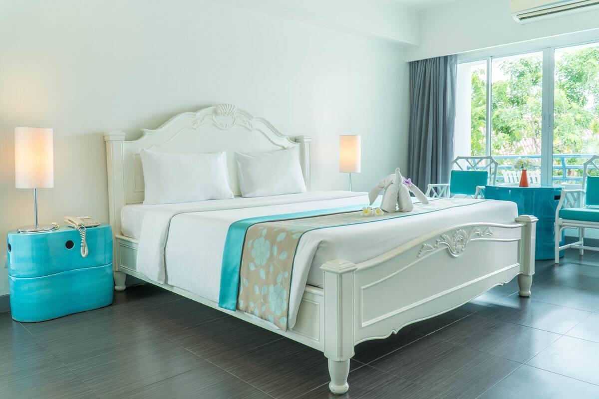Uma cama bem arrumada com cabeceira branca, duas luminárias de apoio e uma mala azul no chão.