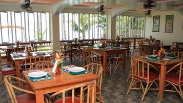 Restaurante com mesas e cadeiras de madeira, janelas amplas e ventiladores de teto.