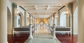 Um corredor longo e estreito com fileiras de bancos brancos e colunas, que leva a uma área central com um candelabro.