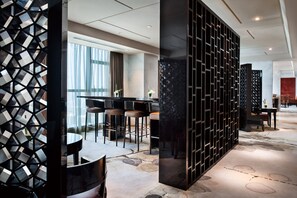 Um lobby de hotel moderno com uma divisória preta geométrica, área de bar com banquetas e piso com padrão de pedra.