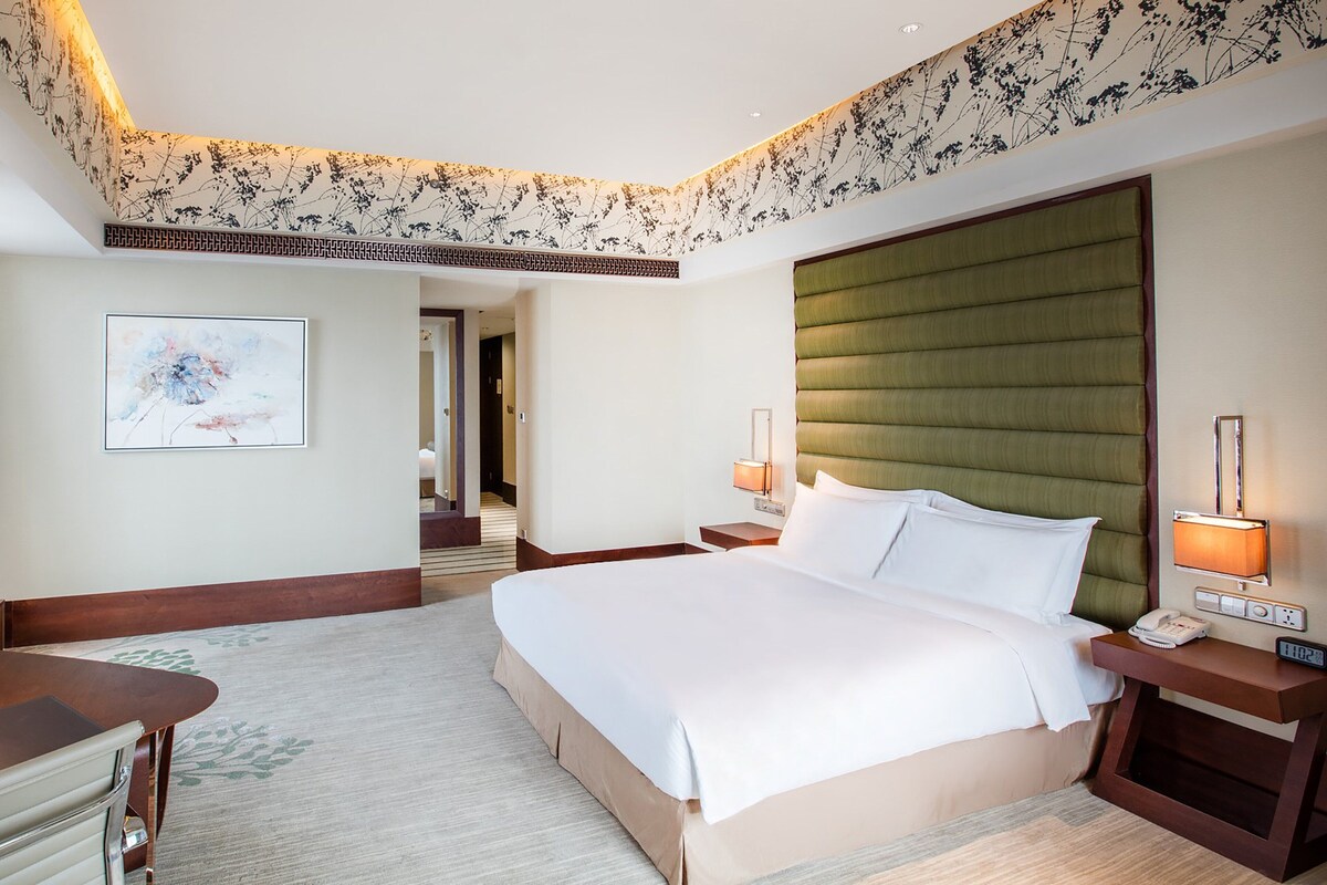 Quarto de hotel com uma cama grande, luminárias de cabeceira, uma escrivaninha e uma cadeira.
