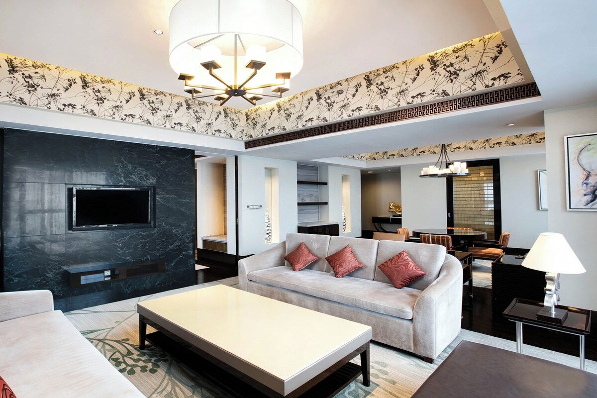 Quarto de hotel moderno com televisão de tela plana, sofá, mesa de centro e área de jantar com lustre.