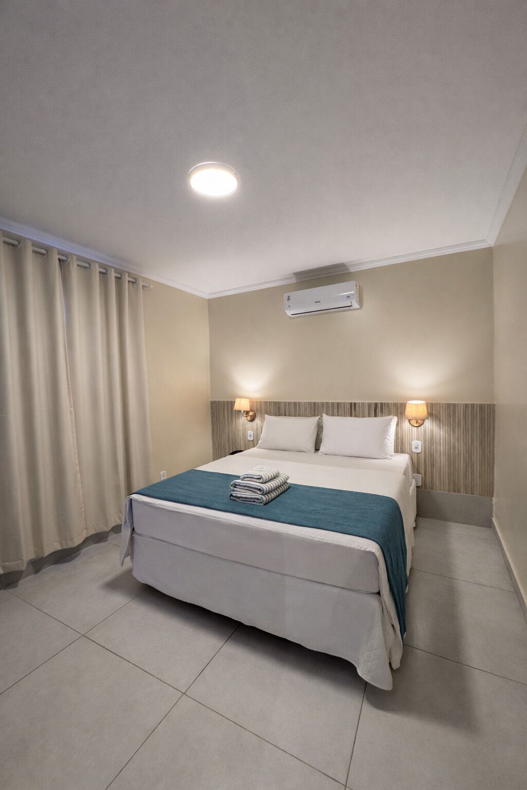 Quarto de hotel com cama, cortinas, ar condicionado e luminárias fixadas na parede.