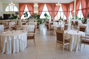 Uma ampla área de jantar com mesas e cadeiras redondas, janelas grandes com cortinas e plantas em vasos.