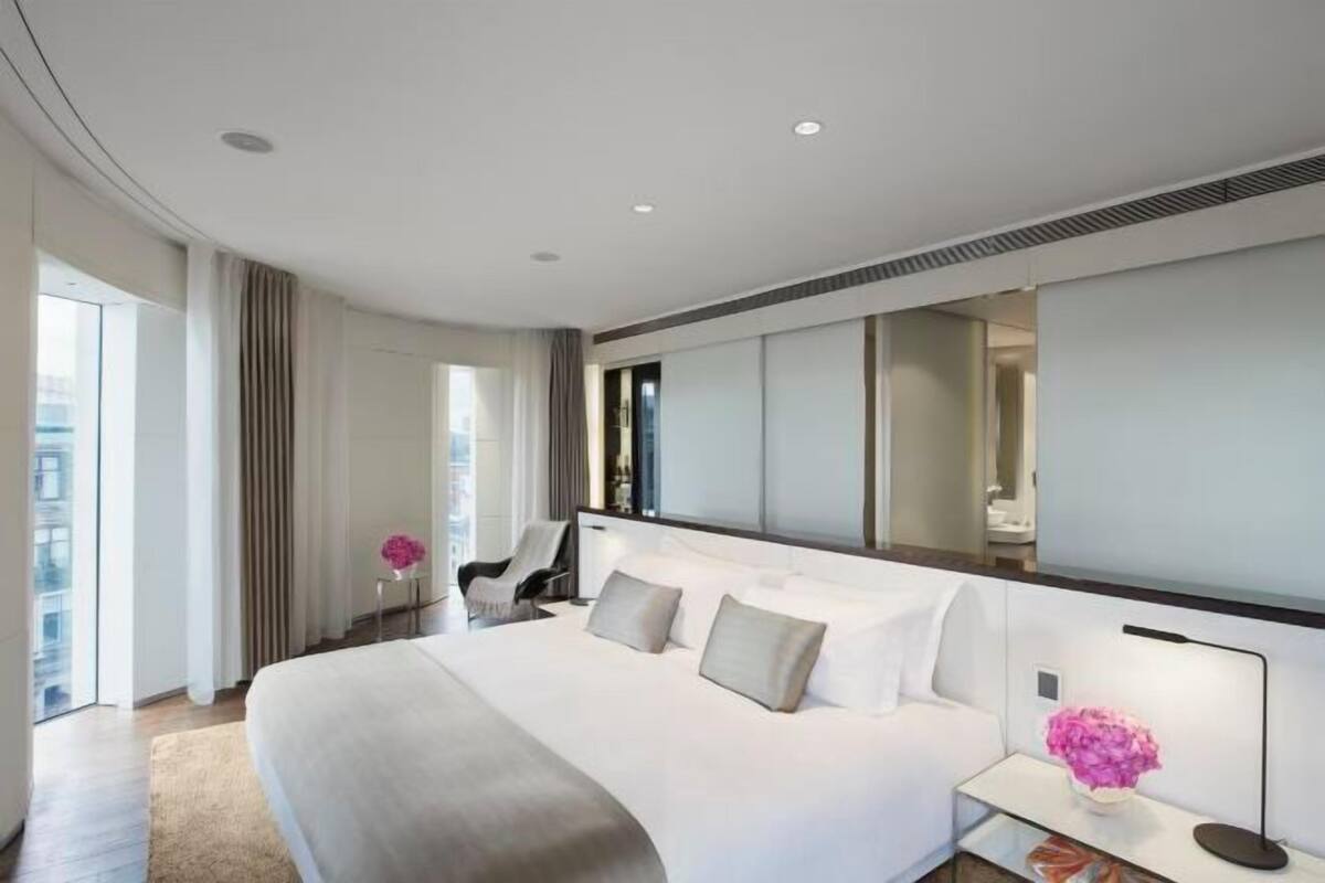 Quarto de hotel moderno com uma cama grande, mesa de cabeceira com abajur, uma cadeira e um espelho amplo.