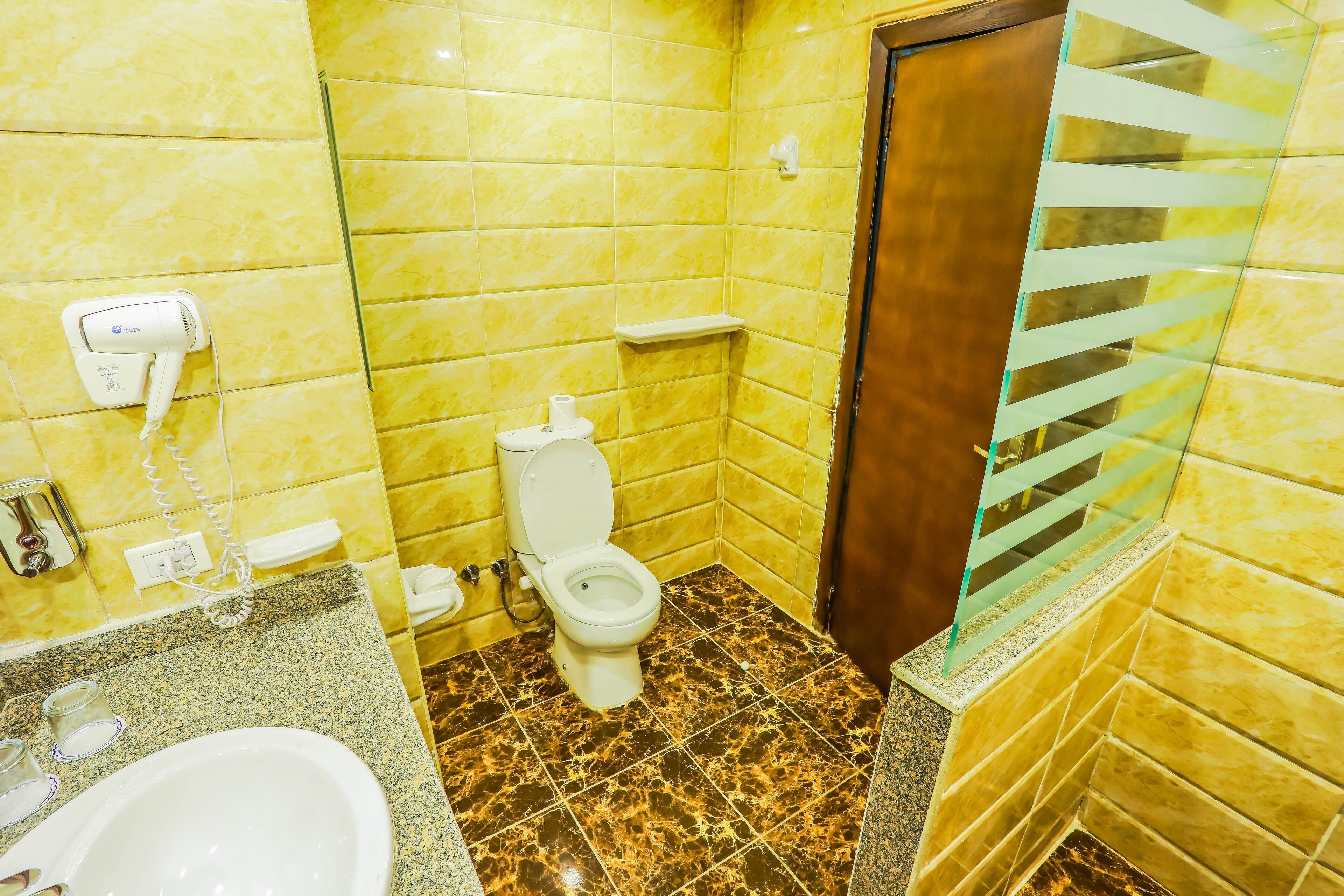 Casa de banho com azulejos amarelos, sanita branca, lavatório com bancada de granito e um secador de cabelo na parede.