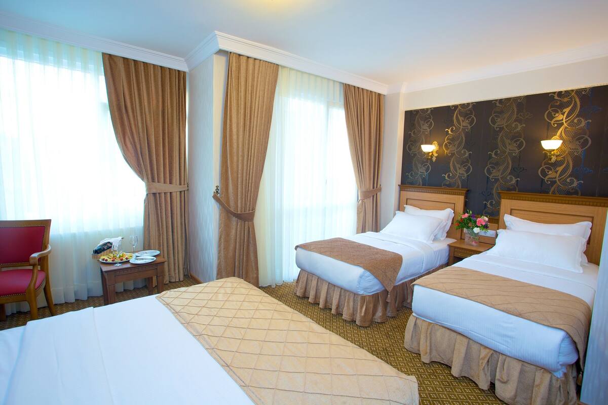 Quarto de hotel com duas camas, uma escrivaninha com cadeira e uma janela com cortinas.