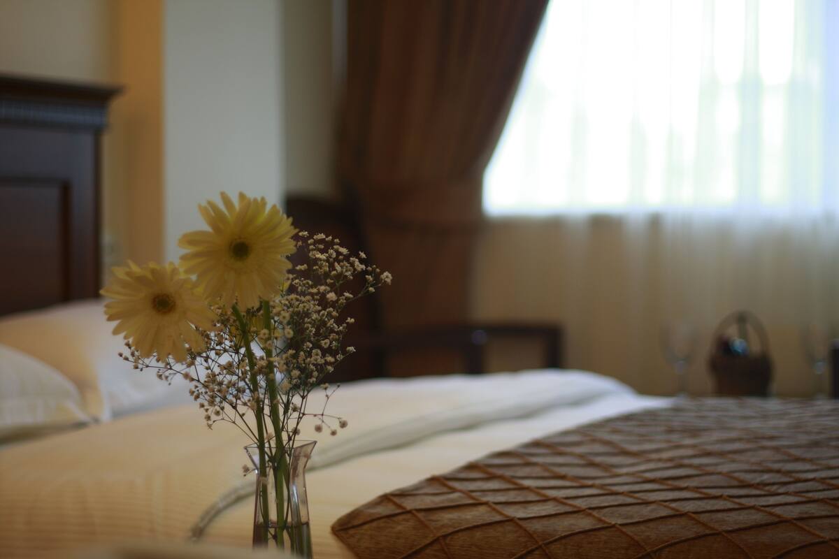Uma cama com cabeceira acolchoada, um vaso com flores e uma janela com cortinas.