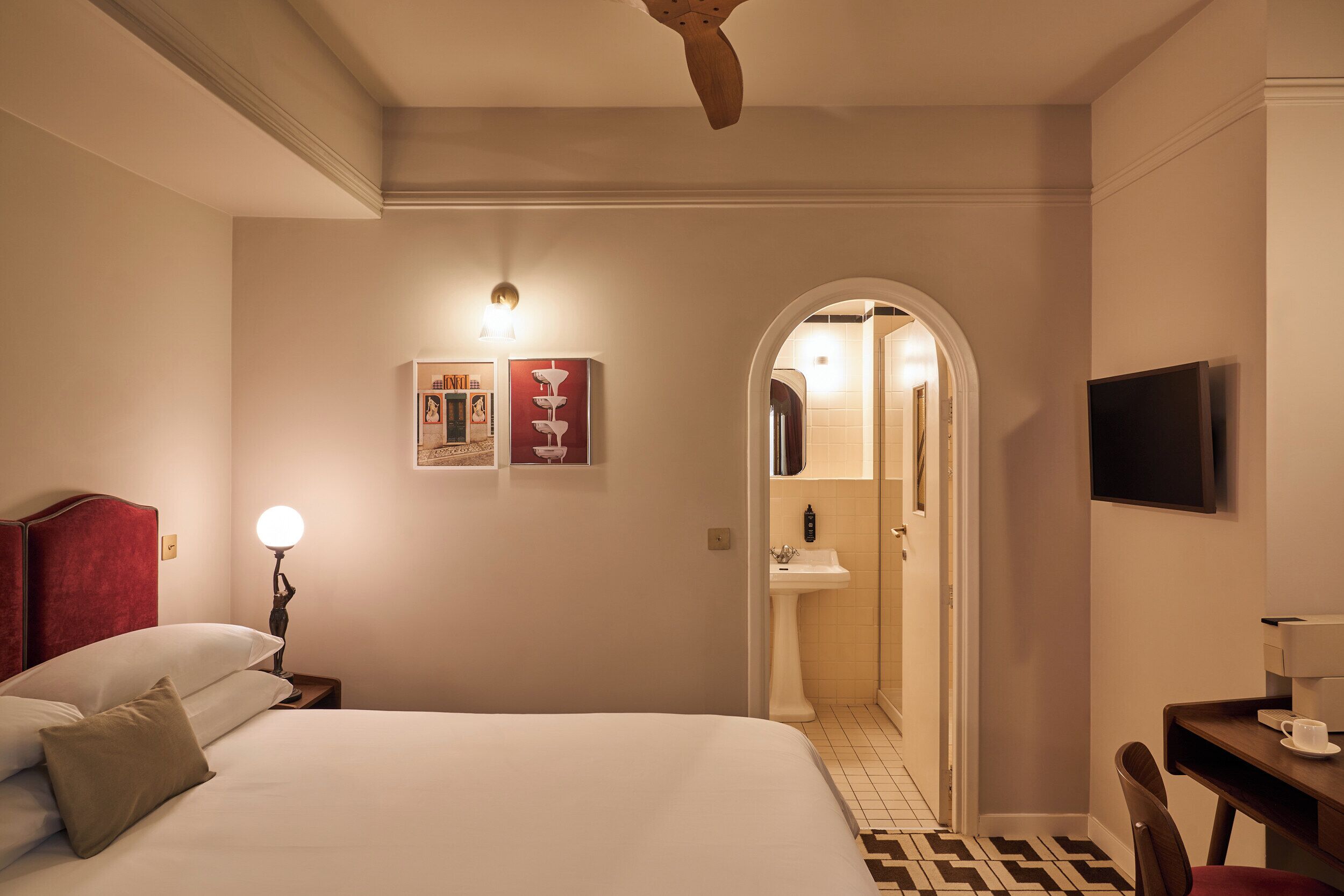 Quarto de hotel com uma cama, uma mesa de trabalho, uma televisão e um banheiro visível por um arco.