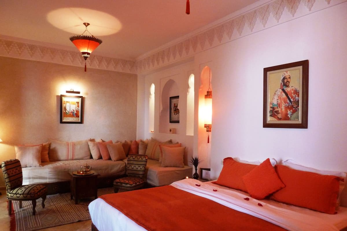 Quarto de hotel com cama, sofá e poltrona, com luminária decorativa no teto e luminárias fixadas na parede.