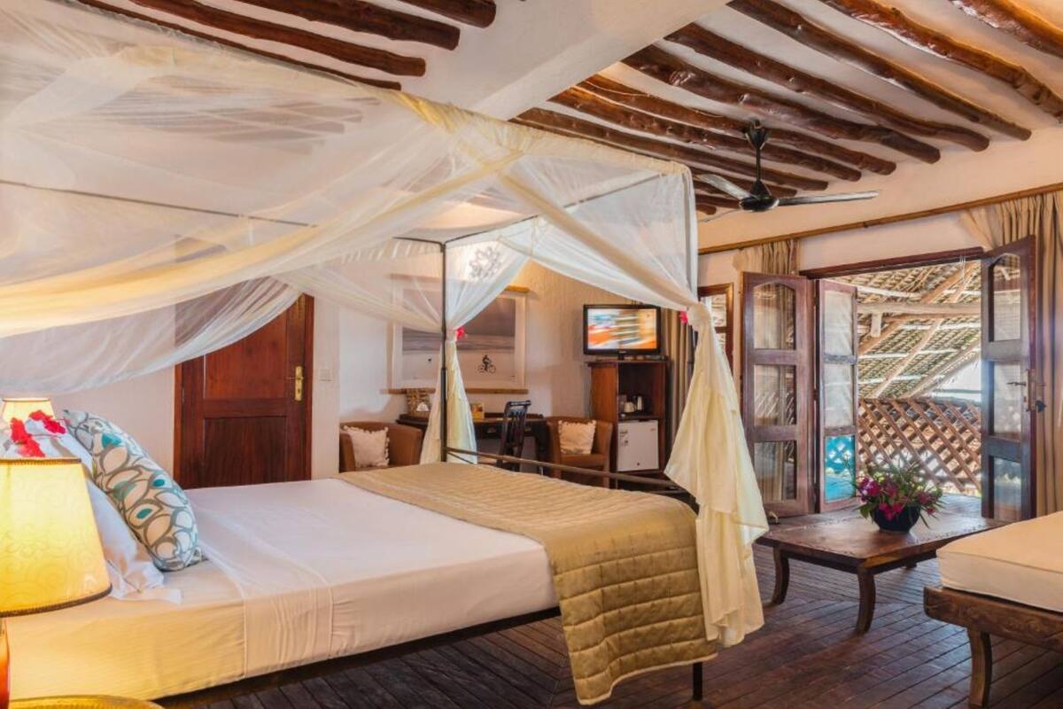Quarto com cama de dossel, piso de madeira, televisão e varanda com grade decorativa.