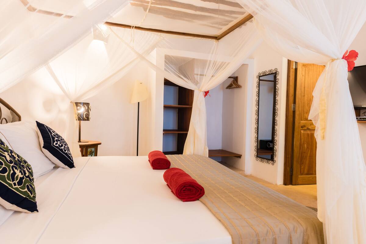 Quarto com cama, mesa de cabeceira, luminária, espelho e porta.