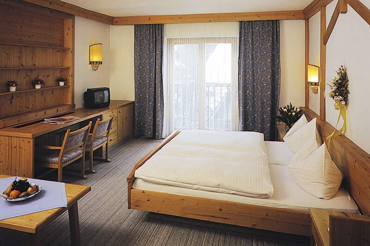 Quarto de hotel com cama de madeira, uma escrivaninha com cadeira, televisão e janela com cortinas.