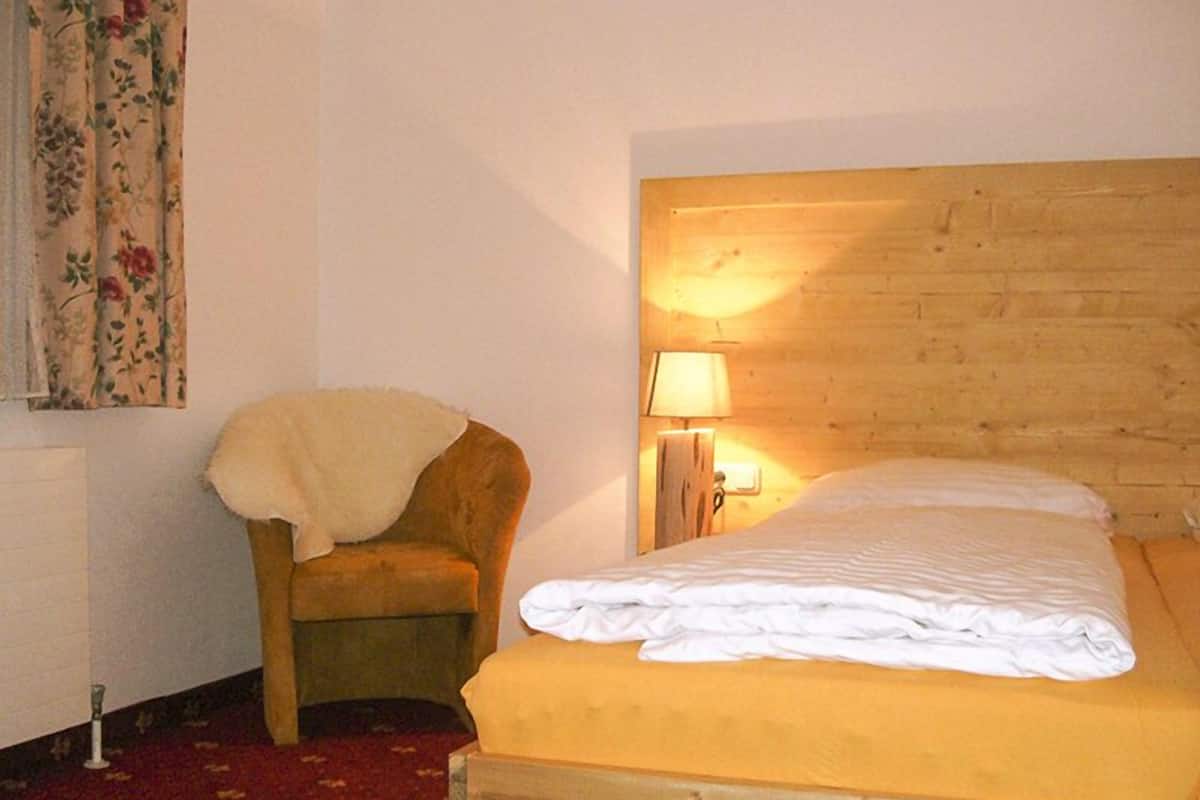 Quarto de hotel com cabeceira de madeira, uma cama de solteiro com roupa de cama branca, uma cadeira com manta bege, um abajur sobre uma mesa de cabeceira de madeira e cortina floral.
