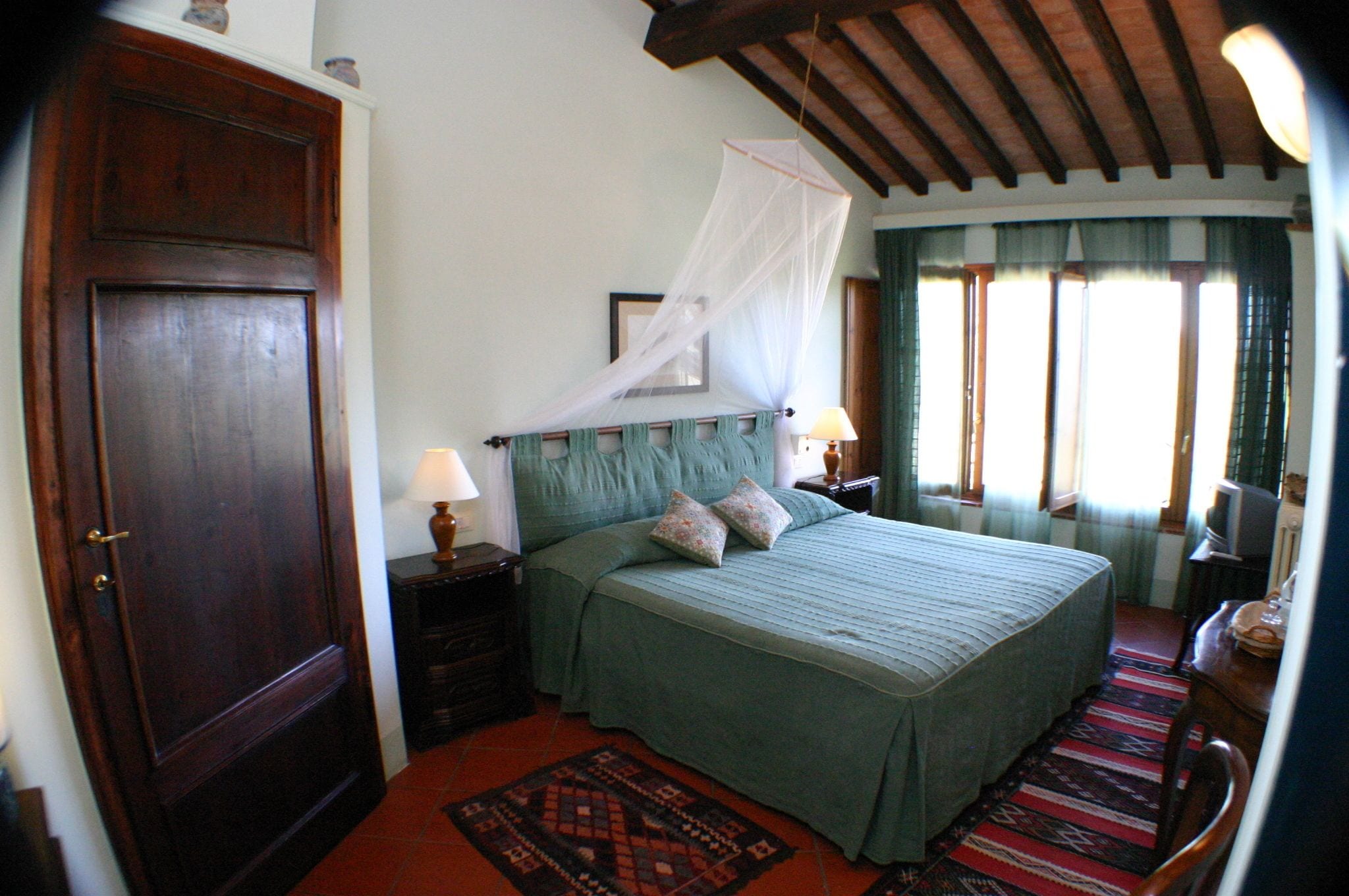 Quarto com uma cama grande, uma porta de madeira, uma mesa de cabeceira com abajur e uma janela com cortinas.