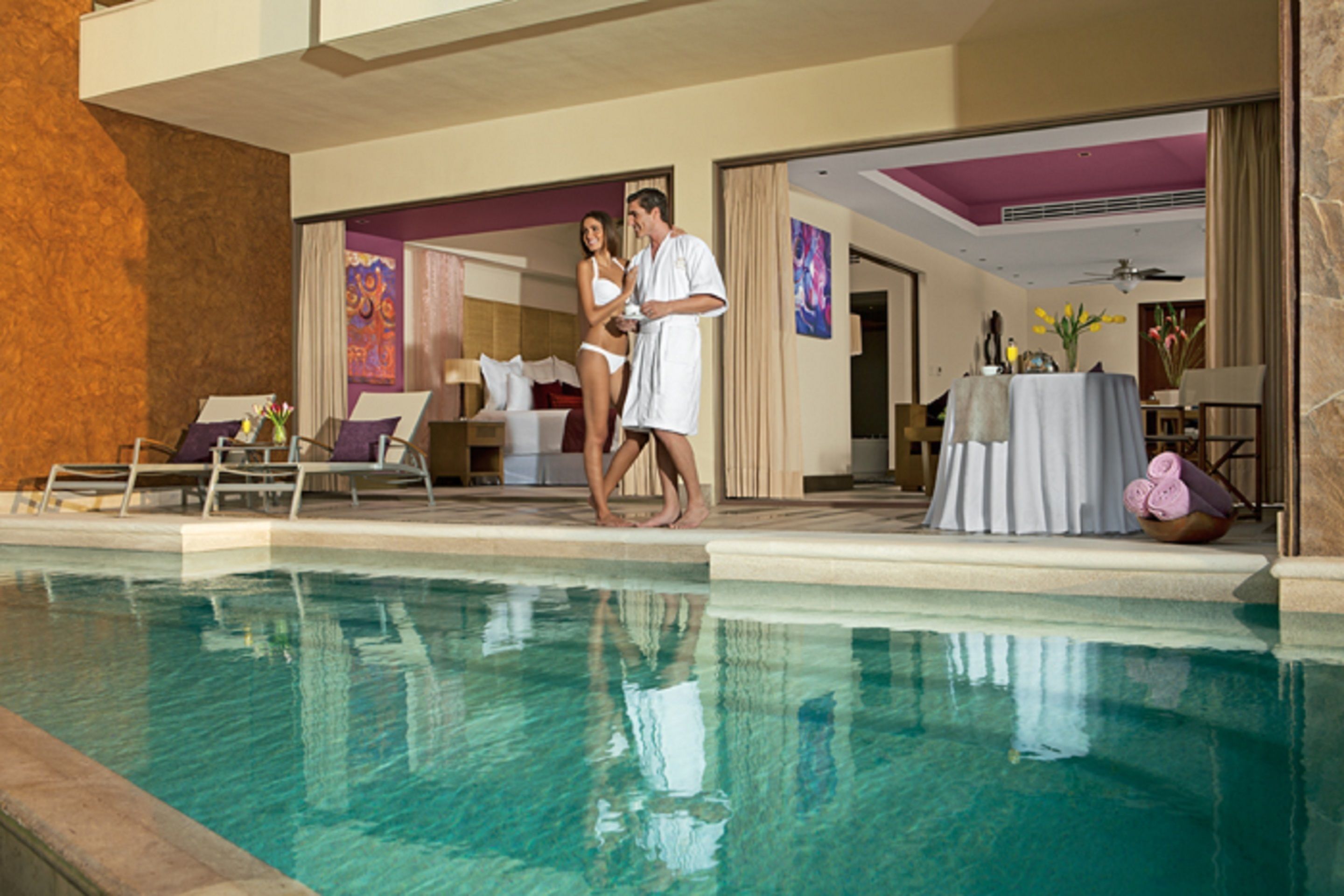 Uma área de piscina de hotel, com um casal ao lado da piscina, um espaço de descanso com cadeiras e uma mesa com toalhas e flores.