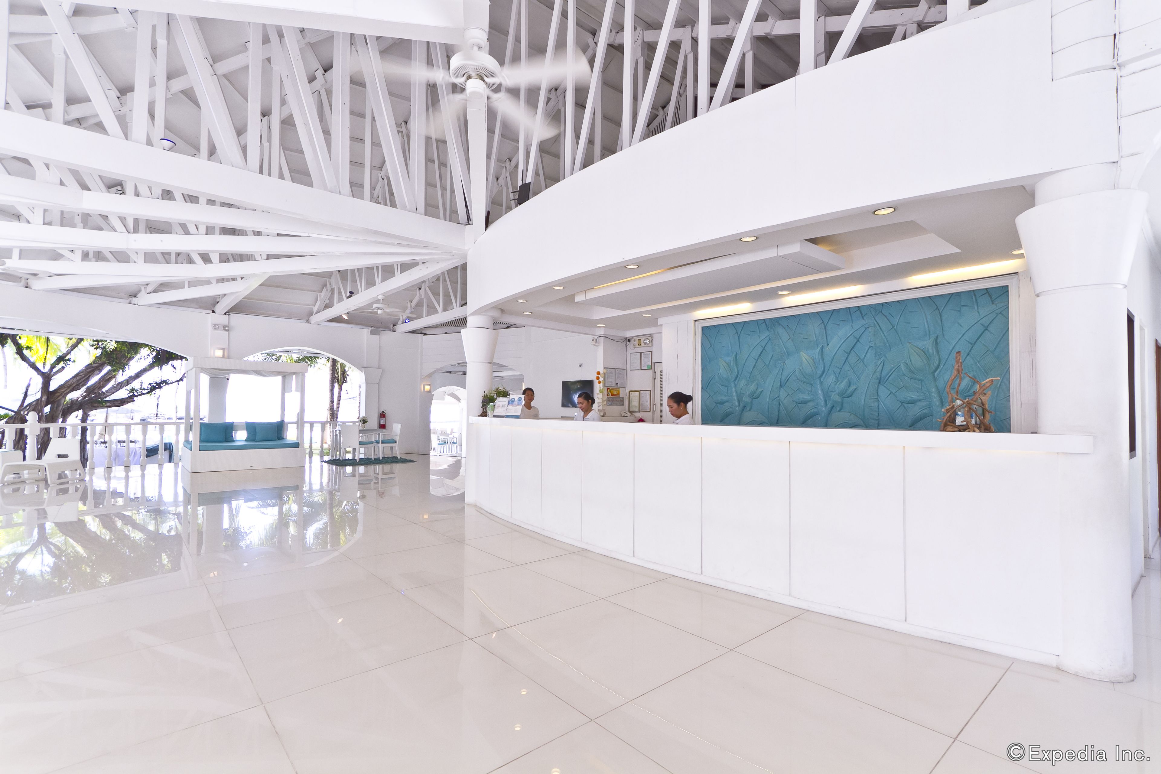 Um hall amplo com uma mesa de recepção, pilares brancos e um teto alto com vigas aparentes.