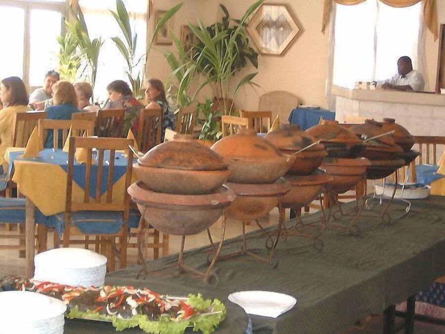 Uma mesa de buffet com várias grandes panelas e recipientes, rodeada por cadeiras e mesas com toalhas azuis.
