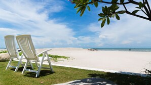 Uma praia com duas cadeiras brancas e uma mesa, céu claro e vista para o oceano.