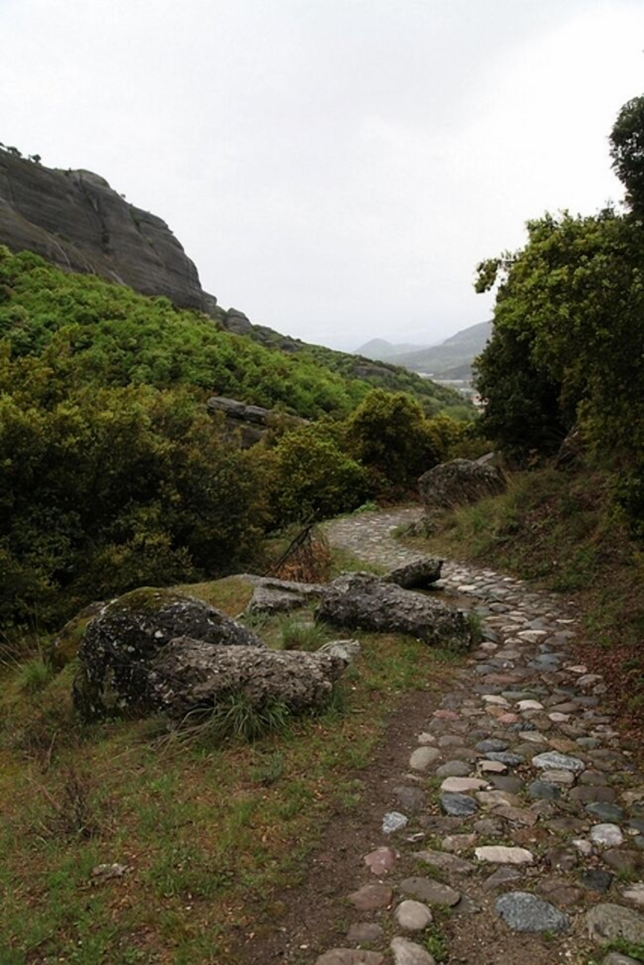 Uma trilha de pedra serpenteia por uma paisagem natural, com vegetação densa e um penhasco rochoso ao fundo.