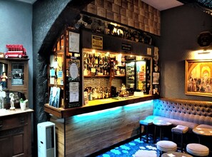 Uma área de bar com balcão de madeira, prateleiras com garrafas e um espaço para sentar com banquetas.