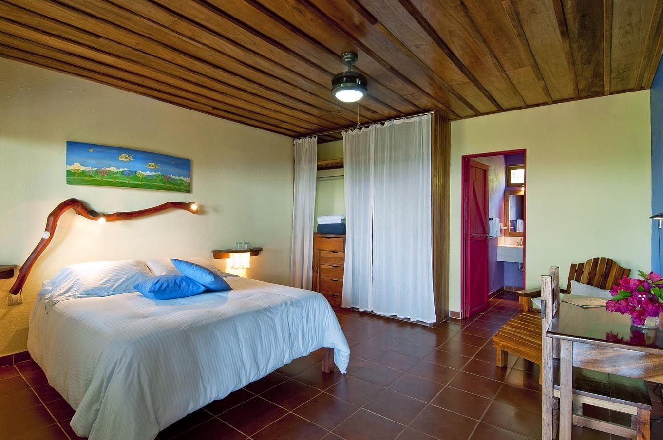 Um quarto com teto de madeira, uma cama com roupa de cama branca e almofadas azuis, um quadro na parede e uma área de refeições com mesa e cadeiras.