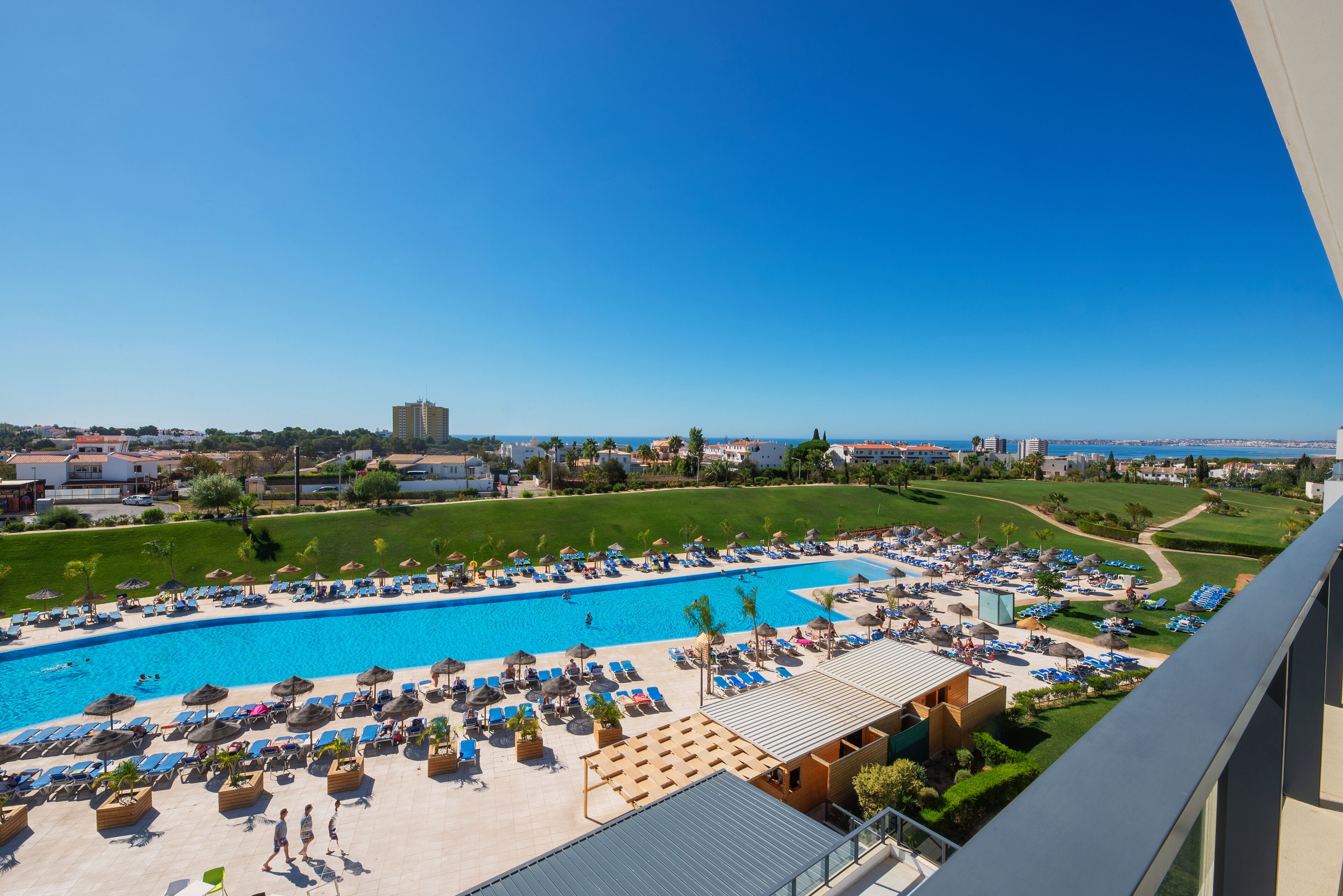 Área de piscina de resort com várias espreguiçadeiras e guarda-sóis, cercada por vegetação e edifícios ao fundo.