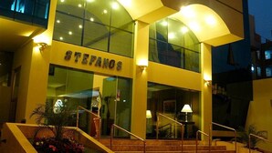 Entrada de hotel com uma placa indicando "STEFANOS", fachada de vidro, escada com corrimãos de metal e uma pequena área de jardim com flores.