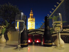 Uma mesa com dois copos de vinho, duas garrafas de Freixenet e duas velas. Um edifício com uma torre é visível ao fundo.