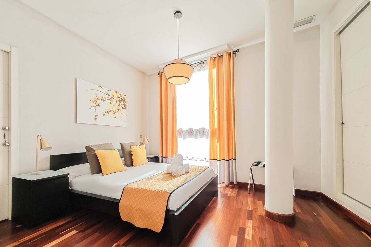Quarto moderno com uma cama grande, piso de madeira e cortinas laranja.