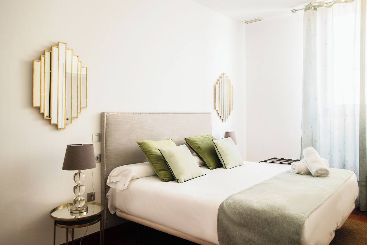 Cama bem arrumada com cabeceira branca, duas almofadas verdes e uma toalha branca ao lado. Há uma pequena mesa com um abajur perto da cama.