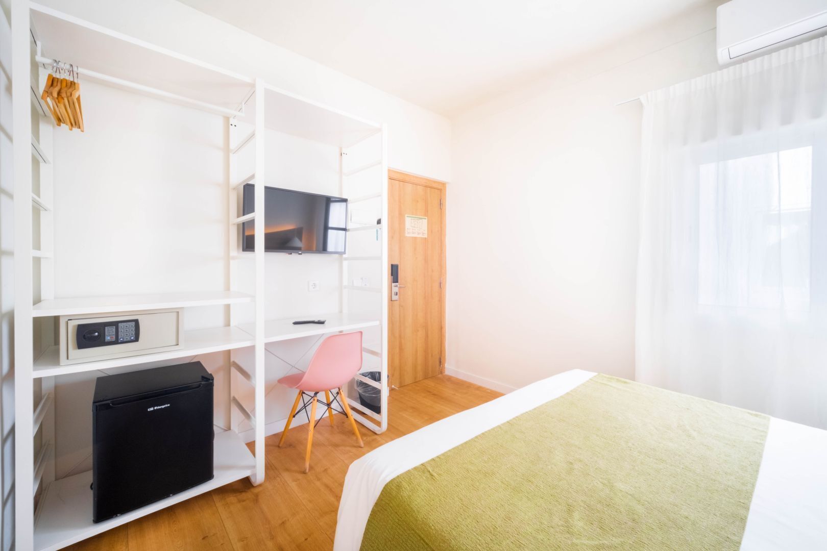 Quarto de hotel com cama, escrivaninha com computador, televisão, guarda-roupa e mini-frigorífico.