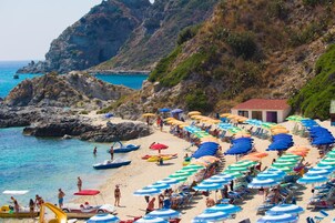 Uma praia com vários guarda-sóis coloridos e espreguiçadeiras, falésias rochosas e um pequeno edifício ao fundo.