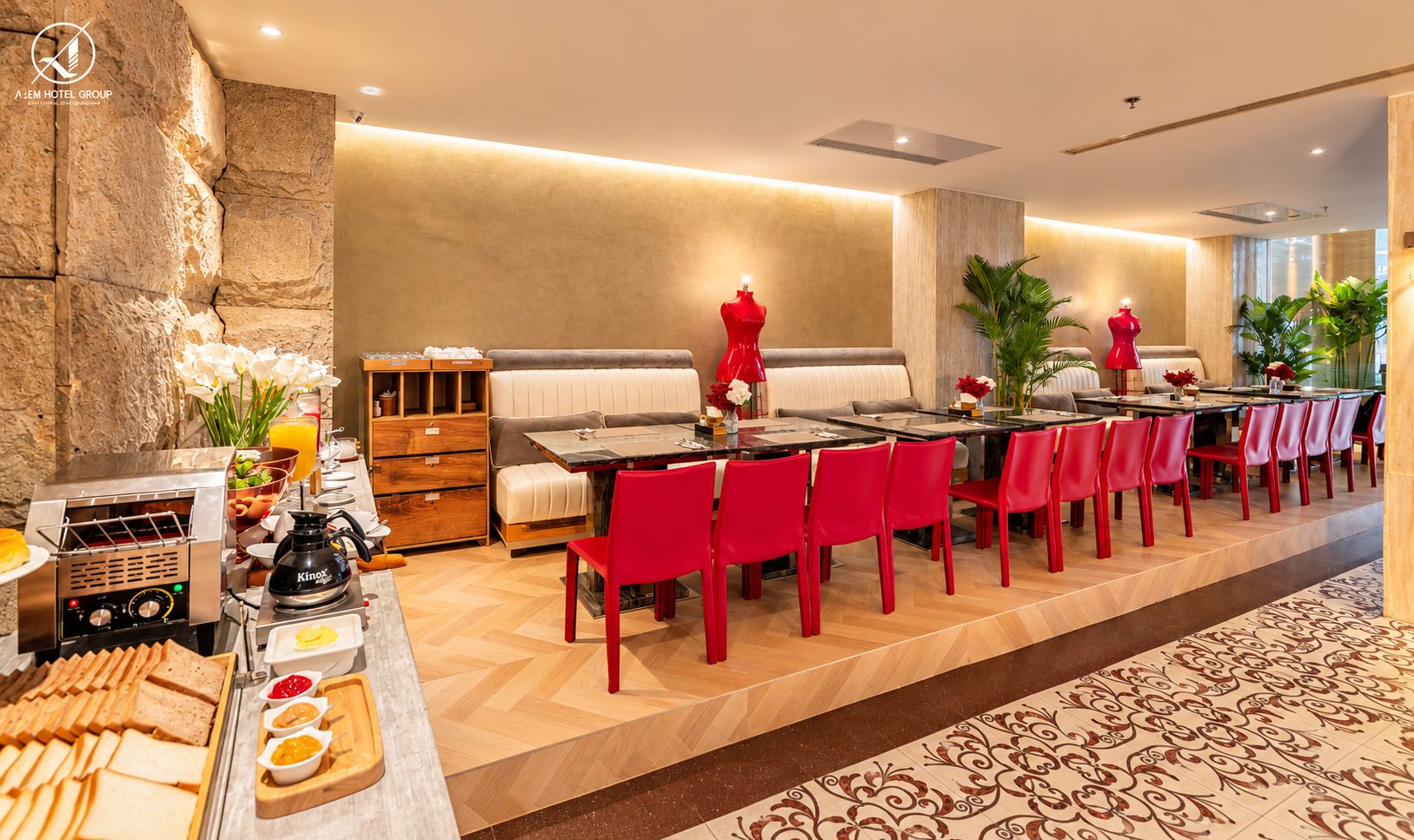 Uma área de refeições de hotel com uma mesa comprida, cadeiras vermelhas, um buffet com pães e patês, e uma máquina de café.