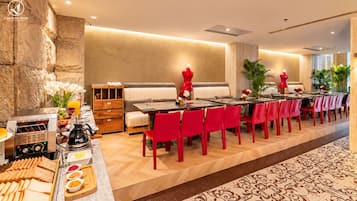 Uma área de refeições de hotel com uma mesa comprida, cadeiras vermelhas, um buffet com pães e patês, e uma máquina de café.