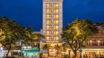 Um hotel com uma placa chamativa, várias janelas e vista para a rua, com carros estacionados e árvores.