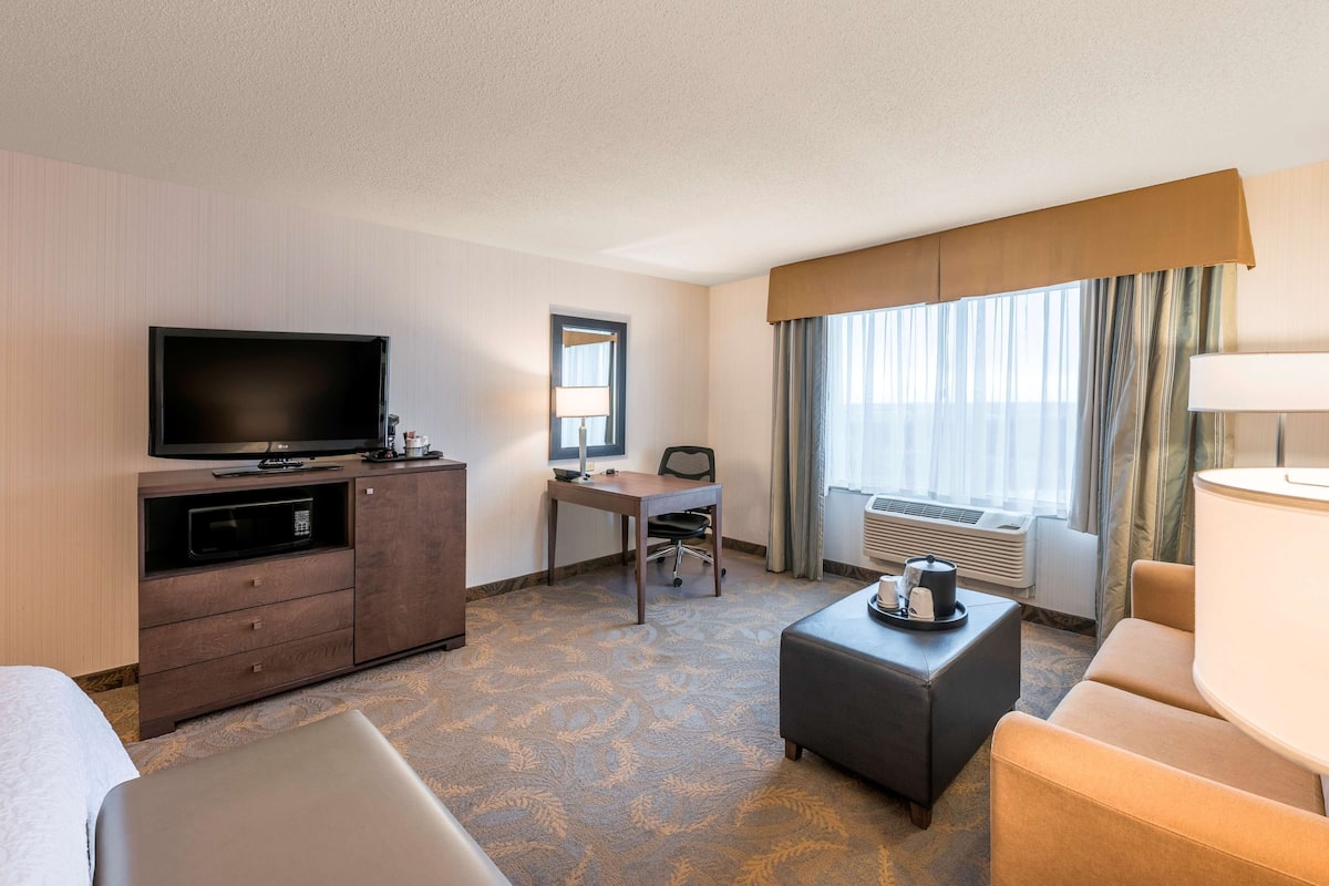 Quarto de hotel com televisão de tela plana, escrivaninha, cadeira, sofá, mesa de centro e janela com cortinas.