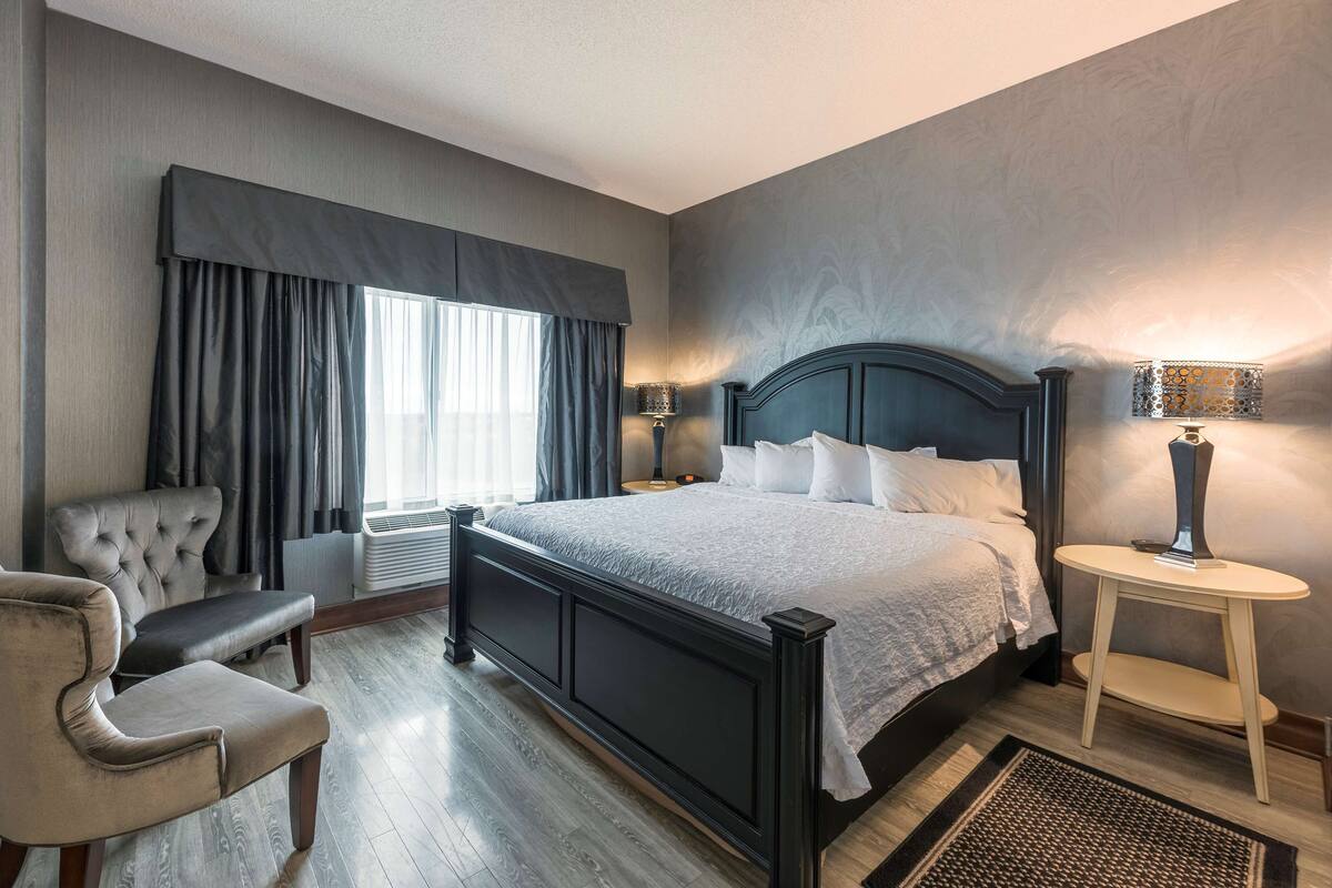 Quarto de hotel com cama grande, uma cadeira, mesa de cabeceira e luminária.