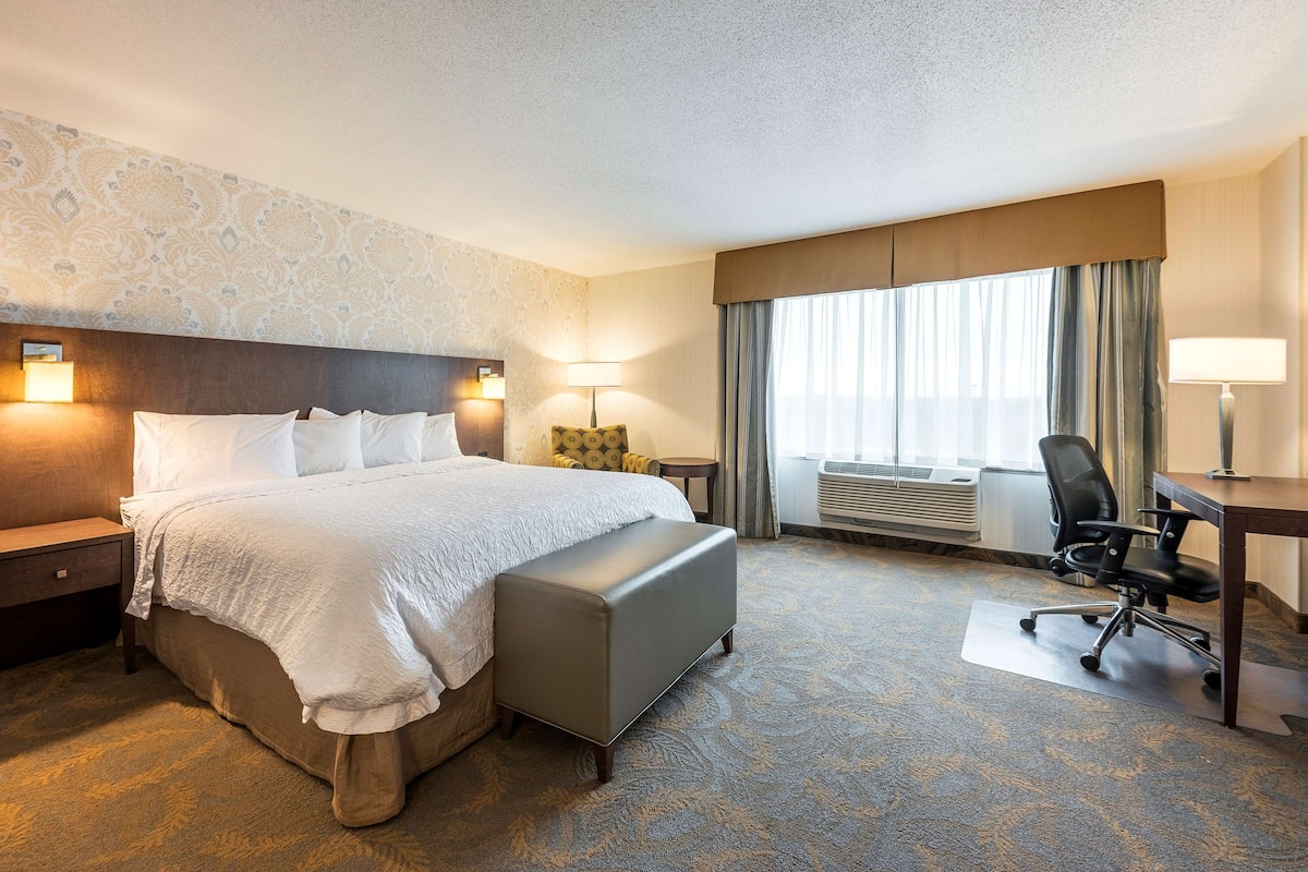 Quarto de hotel com uma cama grande, uma escrivaninha, uma cadeira, um abajur e uma janela com cortinas.