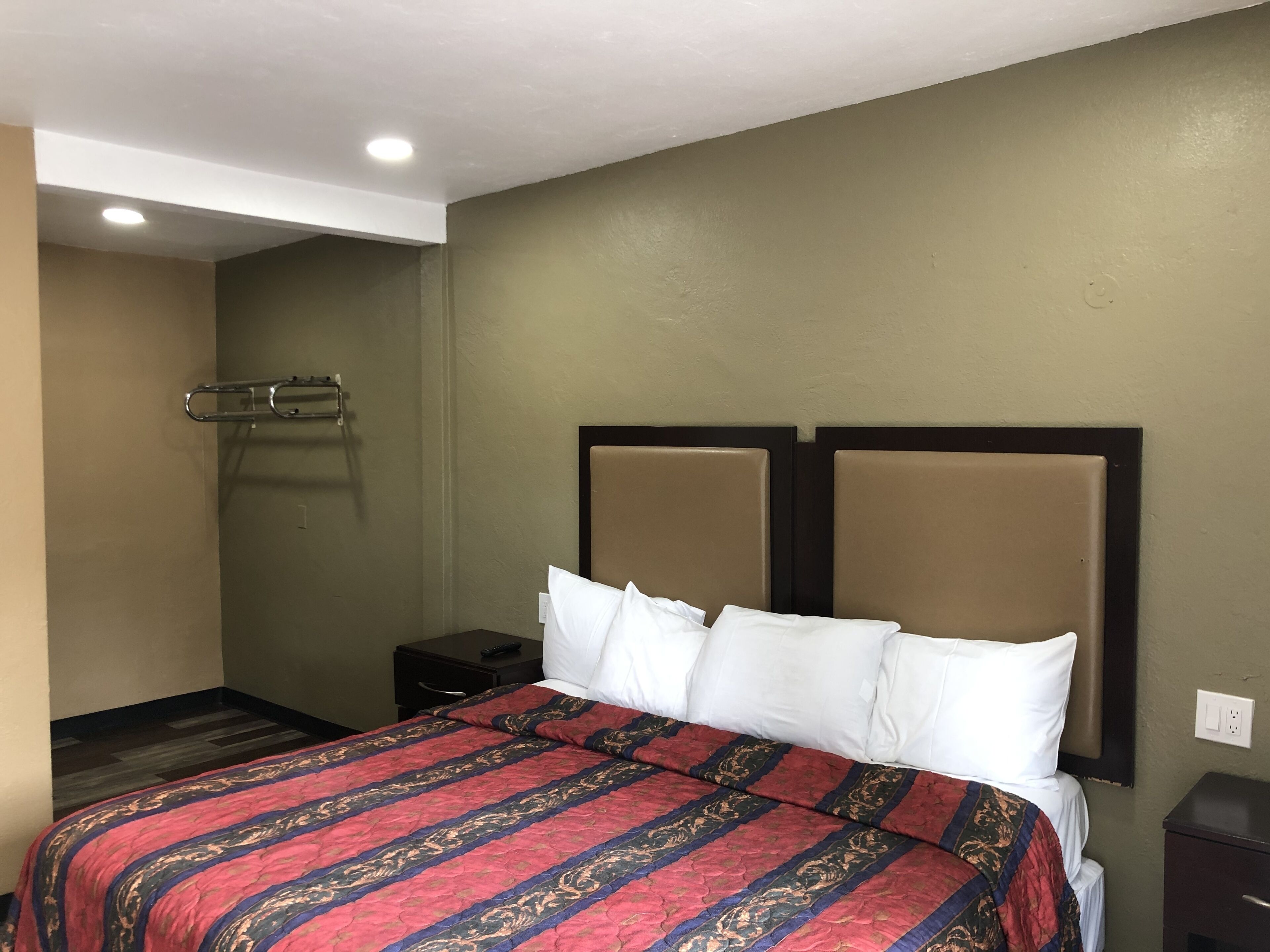 Quarto de hotel com uma cama, uma mesinha de cabeceira, prateleira na parede e uma luminária no teto.