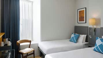 Quarto de hotel com duas camas, uma escrivaninha com cadeira, uma janela com cortinas, uma luminária na parede e um quadro emoldurado.