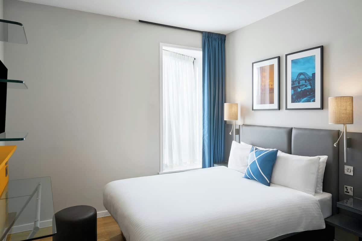 Quarto de hotel moderno com uma cama grande, uma mesa de trabalho e uma janela com cortinas. Há duas fotos emolduradas na parede e um abajur na mesa de cabeceira.