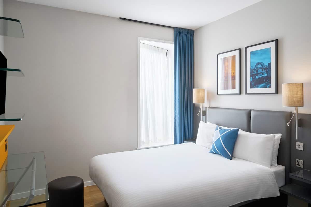 Quarto de hotel com uma cama, uma mesa de cabeceira, uma luminária, uma janela com cortinas e duas fotos emolduradas na parede.