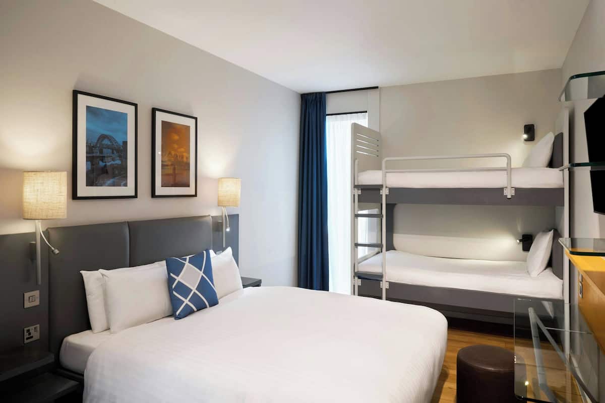 Quarto de hotel com uma cama grande, uma escrivaninha, uma cadeira, uma beliche e duas fotografias emolduradas na parede.