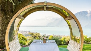 Uma estrutura circular de madeira com uma mesa e bancos, com vista para um lago e montanhas.