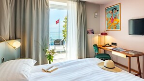 Quarto de hotel com cama, escrivaninha, cadeira, televisão e varanda com uma bandeira.